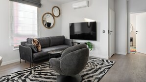 Uma sala de estar moderna com um sofá cinzento, uma poltrona preta, uma televisão de ecrã plano e um tapete estampado.