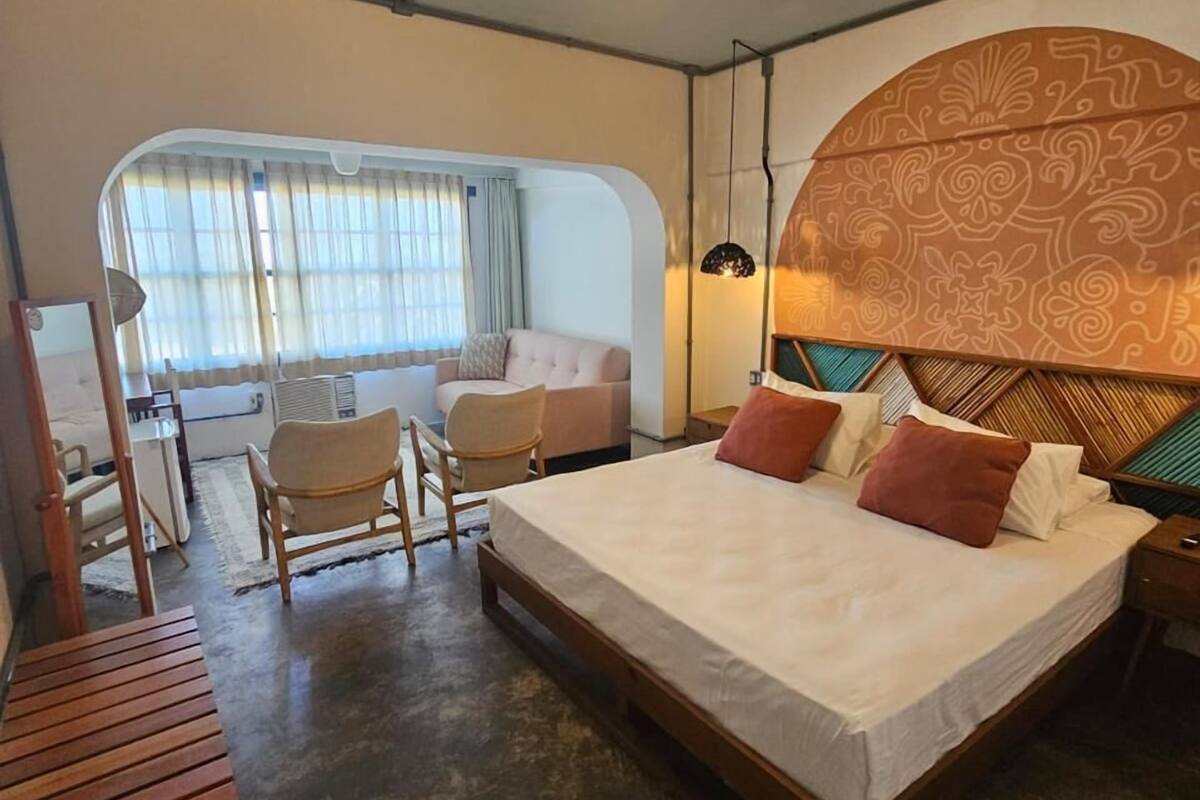 Quarto de hotel com uma cama grande, uma escrivaninha de madeira, uma cadeira, uma janela com cortinas e um cabeceira estampada.