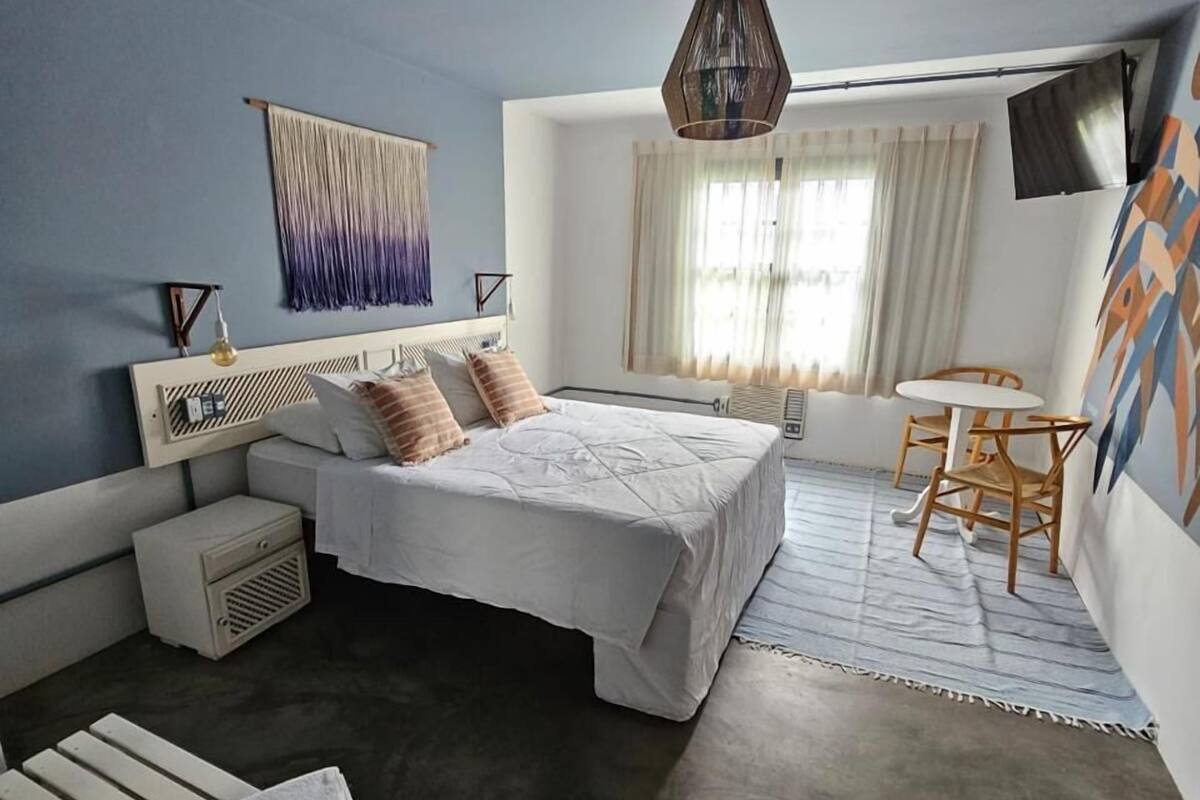 Quarto com cama, mesa de cabeceira, cadeira, televisão e janela com cortinas.