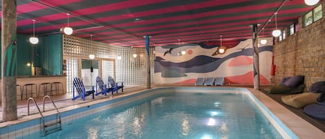 Piscina interna com paredes azulejadas, um mural colorido e cadeiras de descanso.