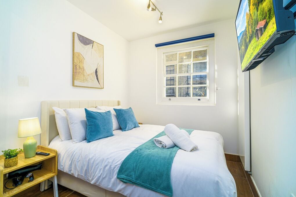 Quarto bem organizado com cama, mesa de cabeceira com luminária, quadro na parede, janela com persianas e televisão fixada na parede.