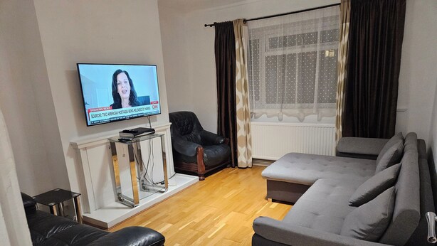 Uma sala de estar com um sofá cinza, uma poltrona de couro preto, uma TV fixada na parede e uma janela com cortinas.