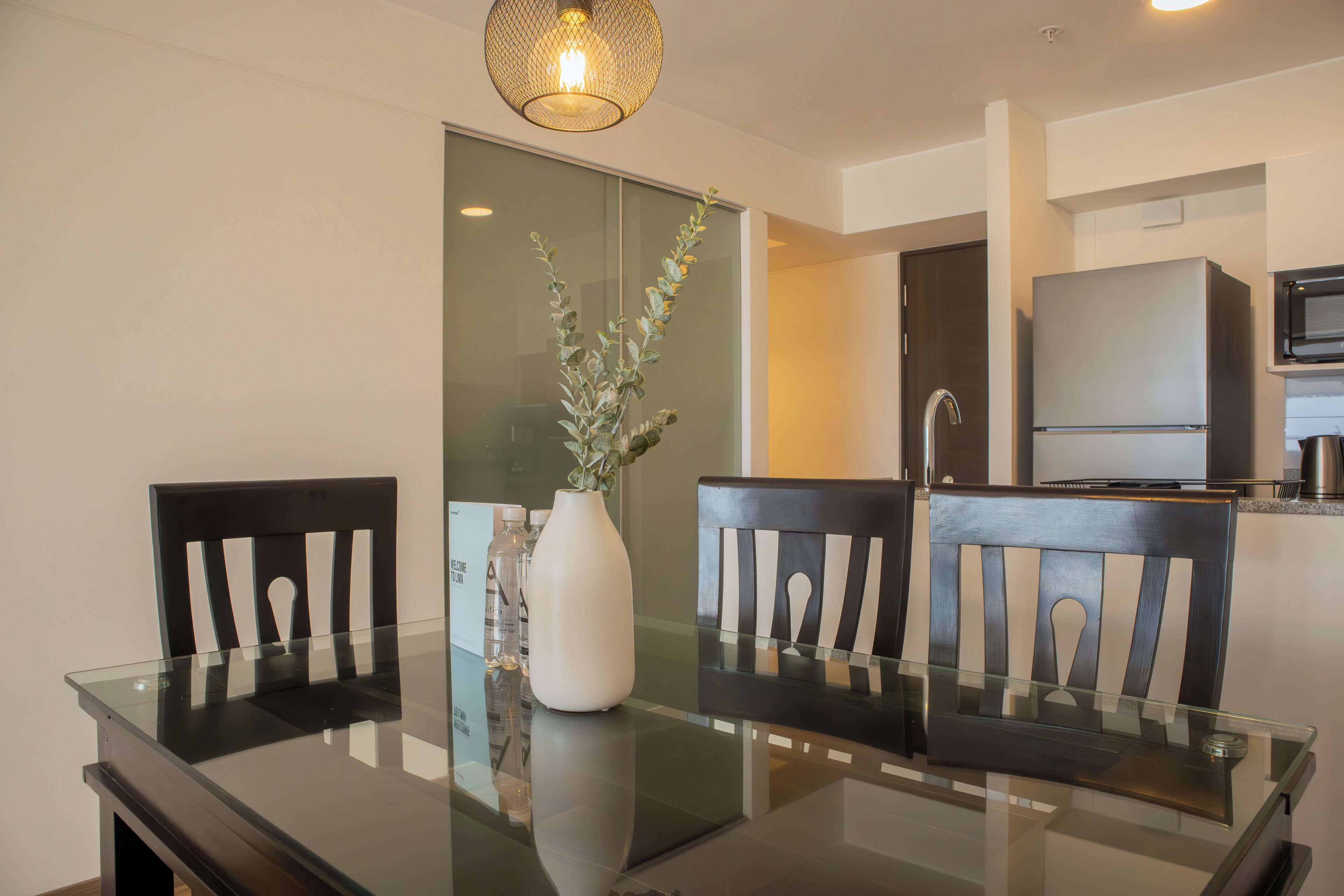 Uma área de jantar moderna com mesa de vidro, duas cadeiras e um vaso com folhagem.