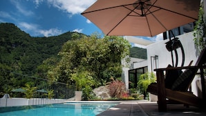Piscina com espreguiçadeira de madeira e guarda-sol, cercada por vegetação exuberante e montanhas ao fundo.