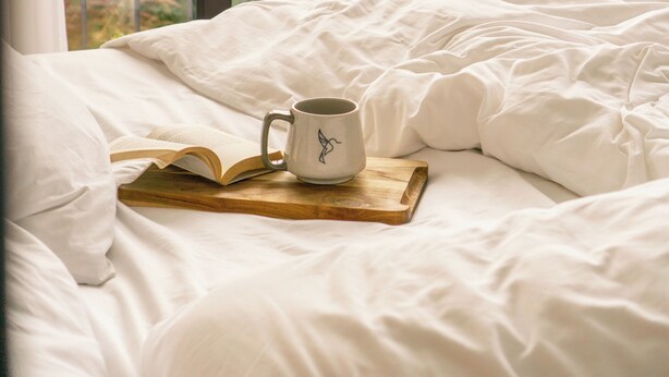 Uma cama com um livro, uma caneca, um tabuleiro de madeira e vista para um lago e montanhas através de amplas janelas.
