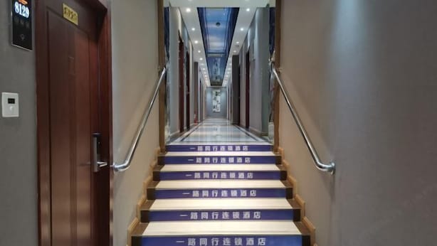 Um corredor de hotel com uma porta de madeira à esquerda, uma escadaria azul e branca com corrimãos e um teto com iluminação embutida.