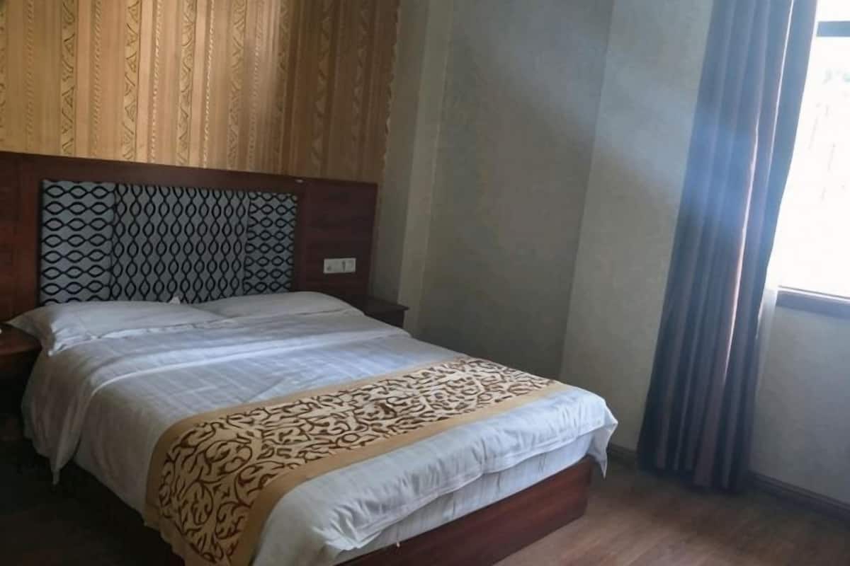 Quarto de hotel com cabeceira de madeira, cama com colcha estampada, mesa de cabeceira e janela com cortinas.