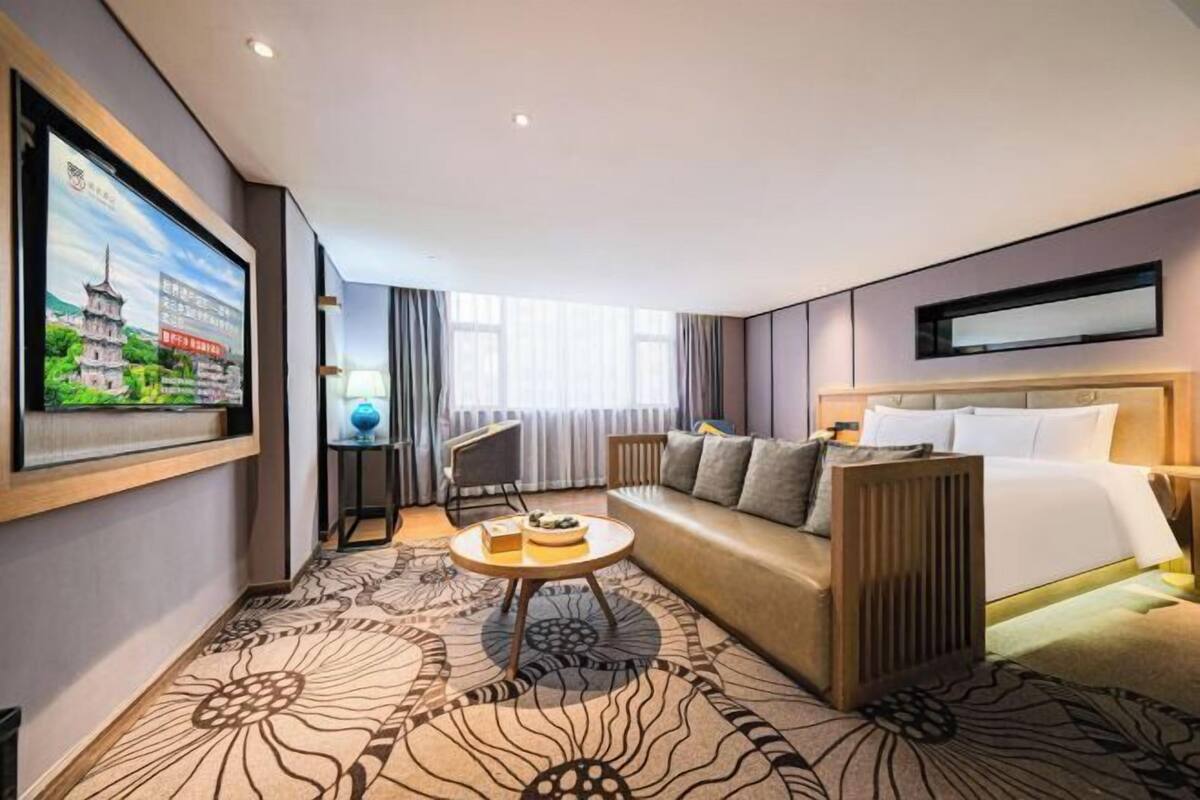 Quarto de hotel com sofá, cama, televisão, mesinha e tapete estampado.