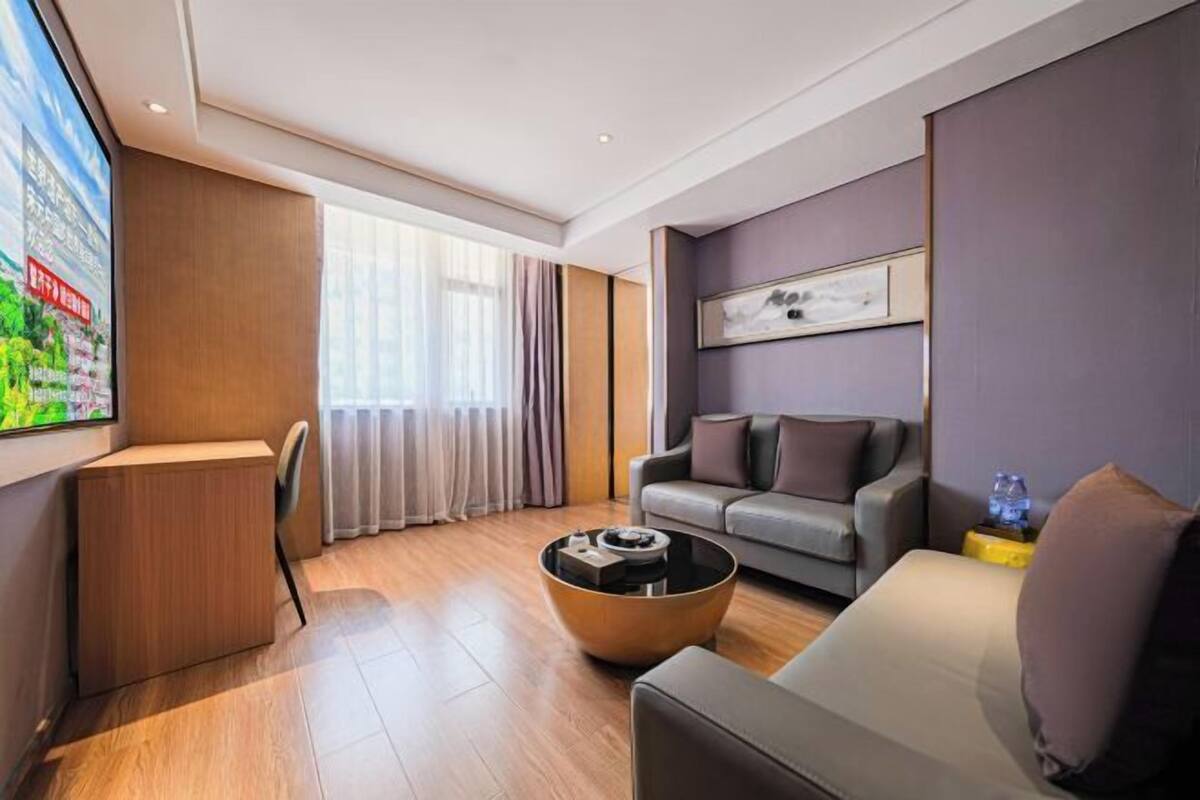 Quarto de hotel moderno com sofá, mesa de centro, televisão e uma grande janela com cortinas.