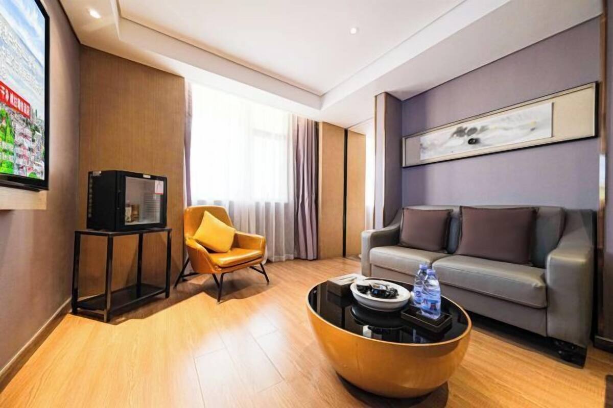 Quarto de hotel moderno com sofá, uma cadeira, mesa de centro, televisão e janela com cortinas.