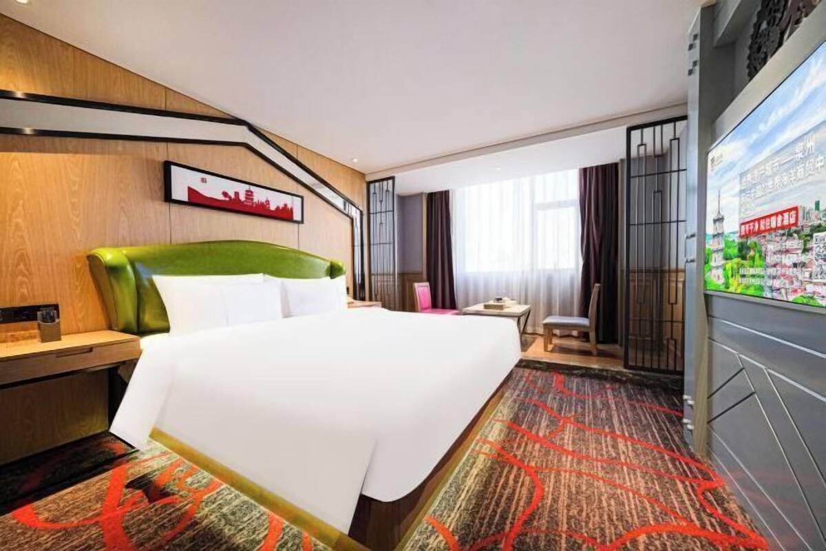 Quarto de hotel com cama, escrivaninha, cadeira, televisão e janela com cortinas.