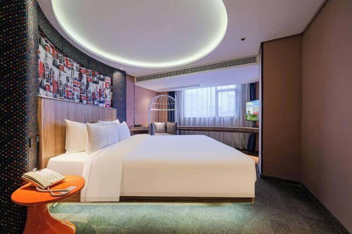 Quarto de hotel com uma cama grande, uma mesinha laranja, um painel decorativo e um candeeiro circular.