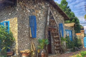 Edifício de pedra com escadaria de madeira que leva a uma porta, rodeado por vasos de plantas.