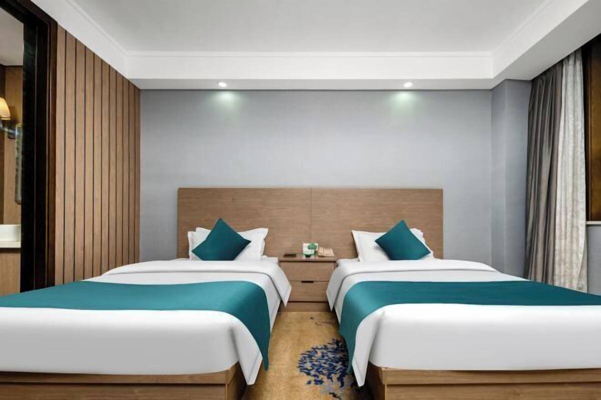 Quarto de hotel com duas camas, uma cabeceira de madeira, mesa de cabeceira e janela com cortinas.
