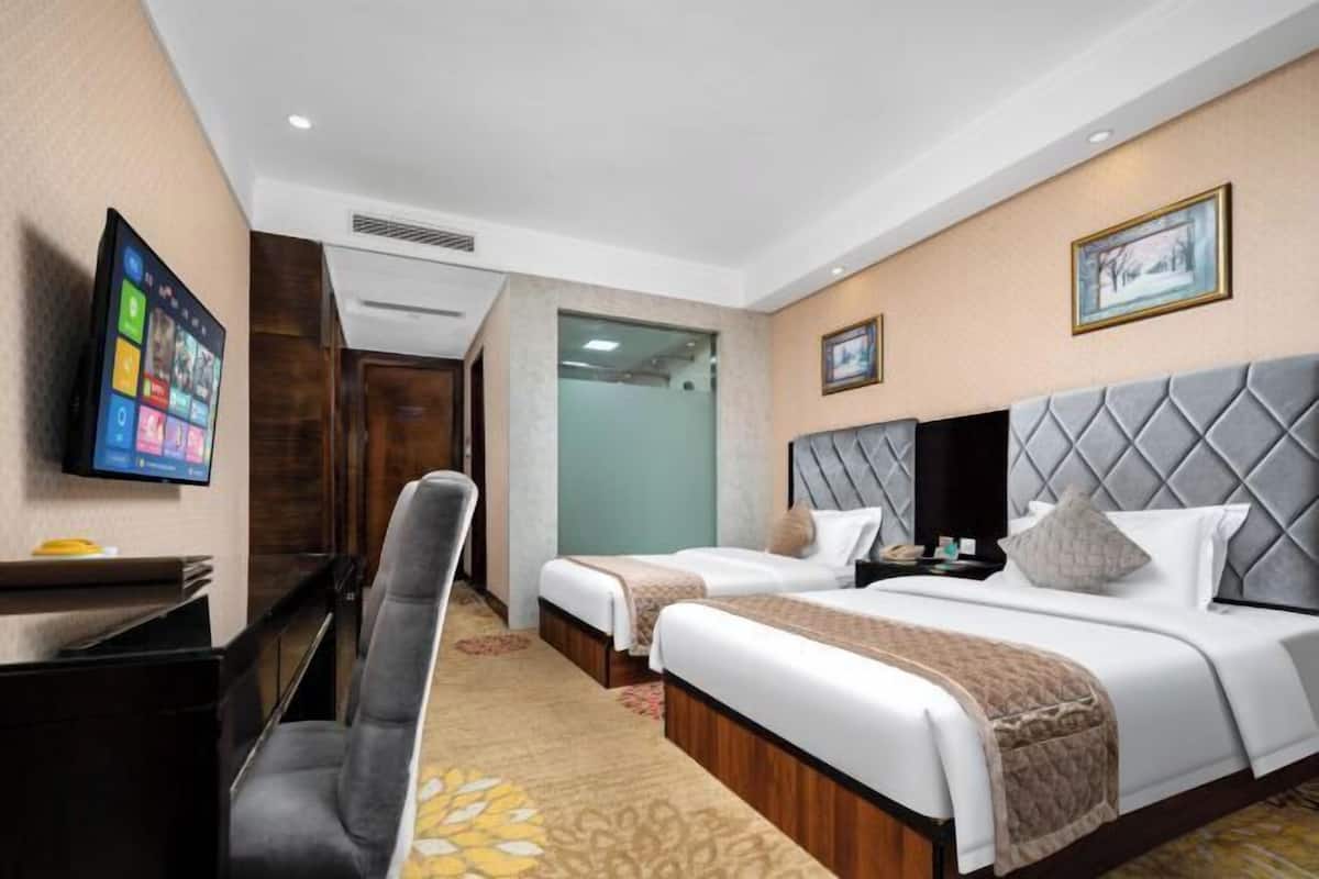 Quarto de hotel com duas camas, uma escrivaninha, uma cadeira, uma televisão e um quadro na parede.