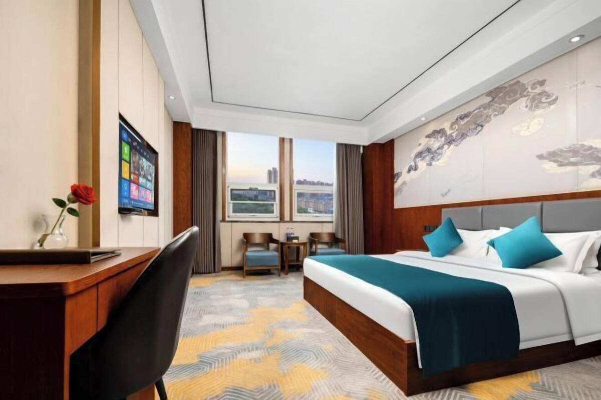 Quarto de hotel com cama, escrivaninha, cadeiras e uma janela grande com vista para a cidade.