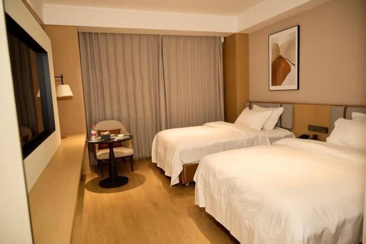 Quarto de hotel com duas camas, uma escrivaninha, uma cadeira e uma janela grande com cortinas.