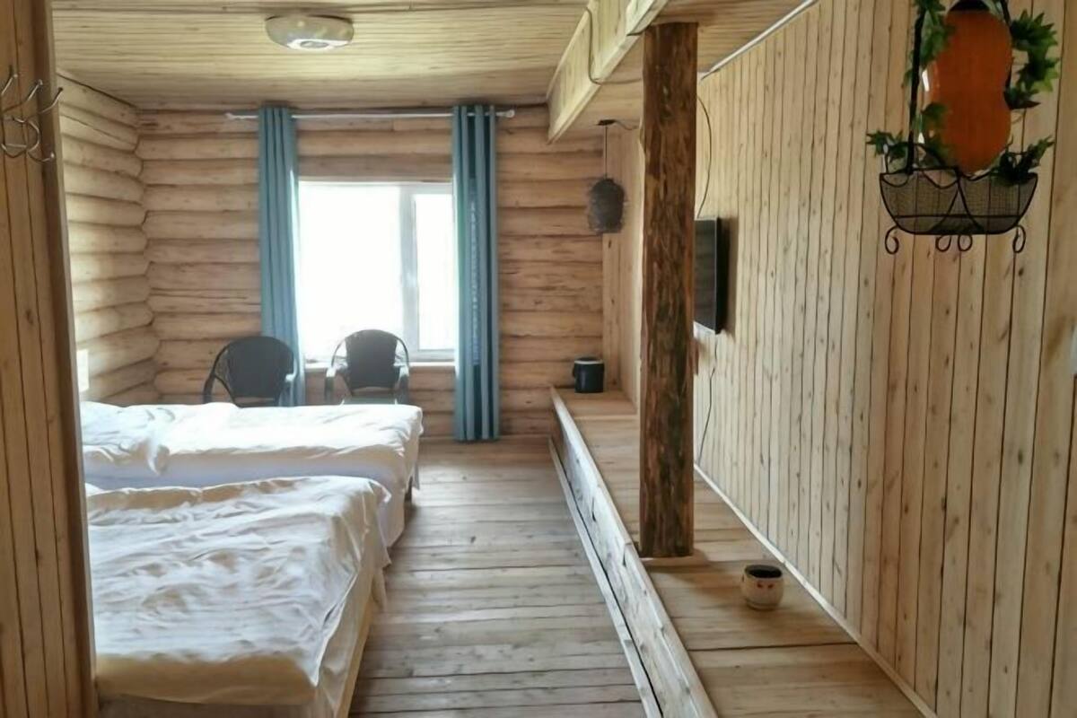 Quarto de cabana de toras com duas camas, uma cadeira, uma TV e uma janela com cortinas.