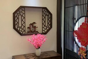 Espelho fixado na parede com design geométrico, uma pequena mesa com um vaso de flores cor-de-rosa e um objeto decorativo sobre a mesa.