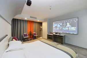 Quarto de hotel moderno com uma cama grande, uma TV exibindo uma videoconferência, uma mesinha com cadeiras e uma planta decorativa.