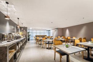 Uma área de refeições moderna com várias mesas e cadeiras, um buffet e uma grande janela que proporciona luz natural.