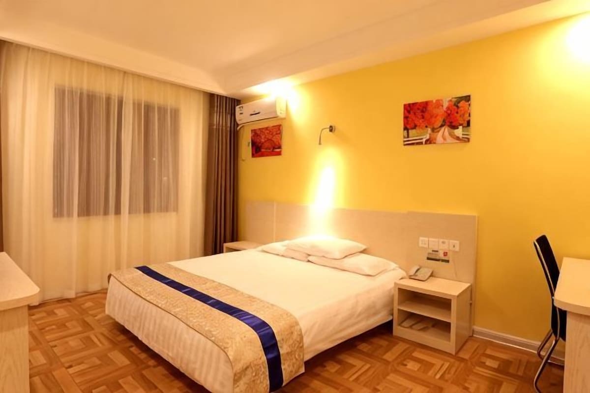 Quarto de hotel com cama, mesa de cabeceira, cadeira, escrivaninha, luminária e janela com cortinas.