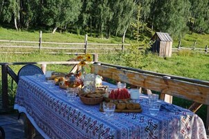 Mesa de refeições ao ar livre com comida e bebida, rodeada por uma cerca de madeira e vegetação.