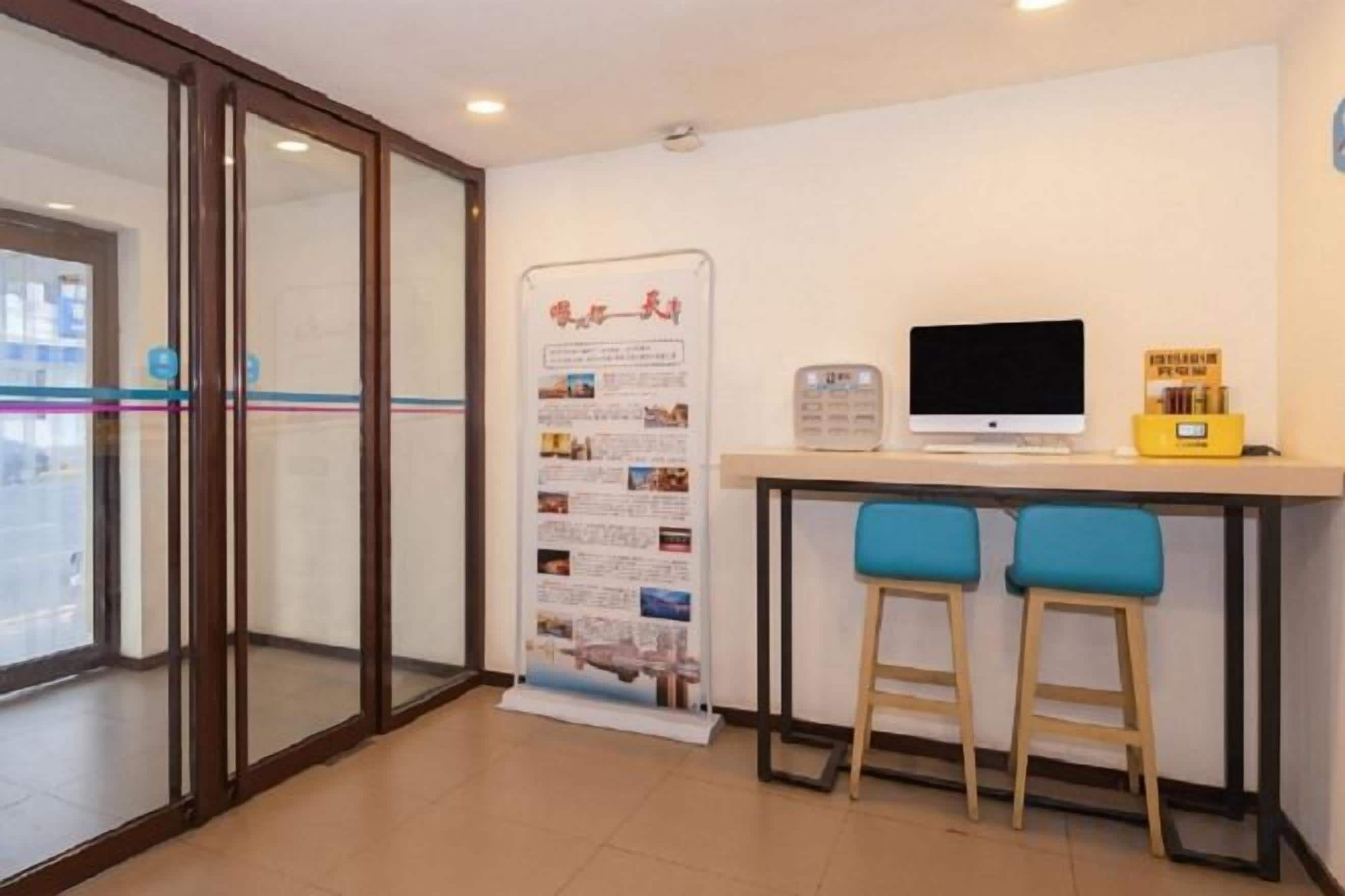 Uma área de recepção com entrada por porta de vidro, um monitor de computador, uma pequena mesa e dois banquetas azuis.