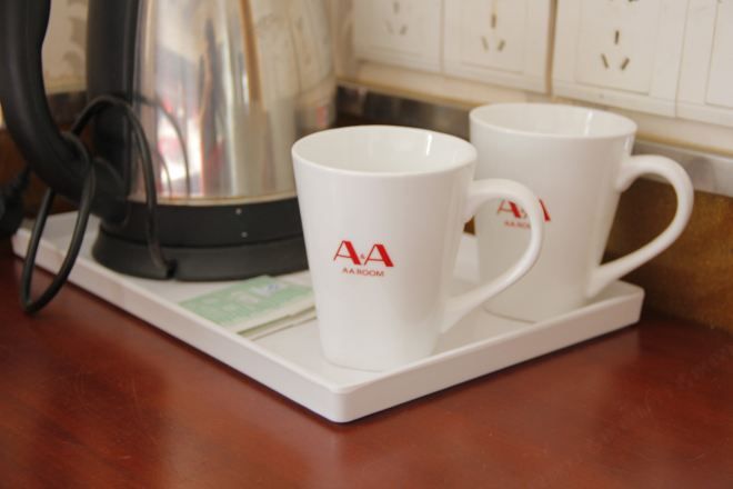 Duas canecas brancas com o logotipo 'AA ROOM' sobre uma bandeja branca, colocadas numa superfície de madeira com uma máquina de café ao fundo.