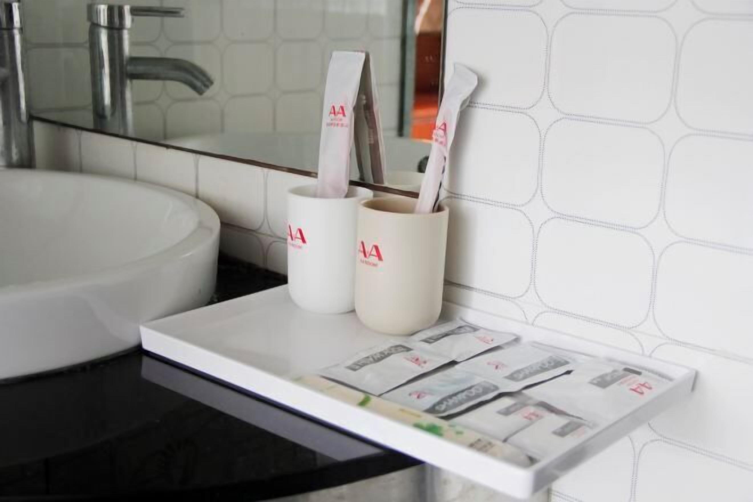 Um banheiro com pia branca, dois dispensadores brancos com logotipos vermelhos e um porta-revistas com diversos itens.