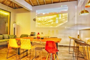 Sala de estar moderna com mesa de jantar de madeira, banquetas de bar e uma tela grande exibindo uma projeção de paisagem.