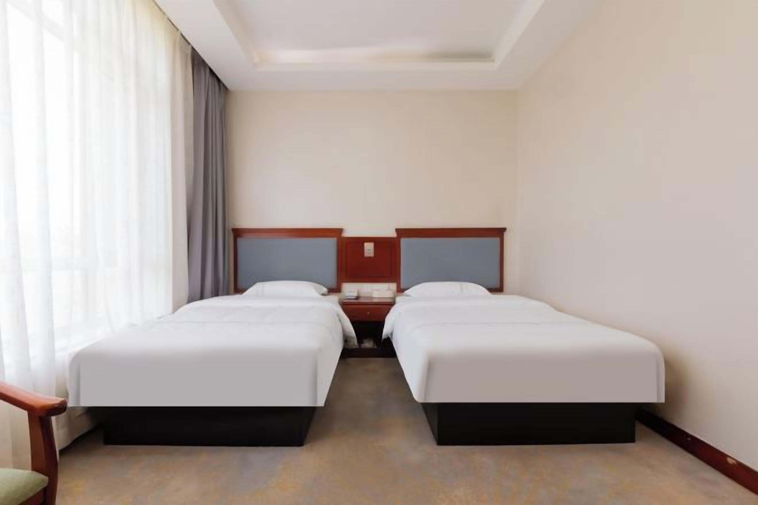 Quarto de hotel com duas camas de solteiro, cabeceira de madeira, mesa de cabeceira e janela com cortinas.
