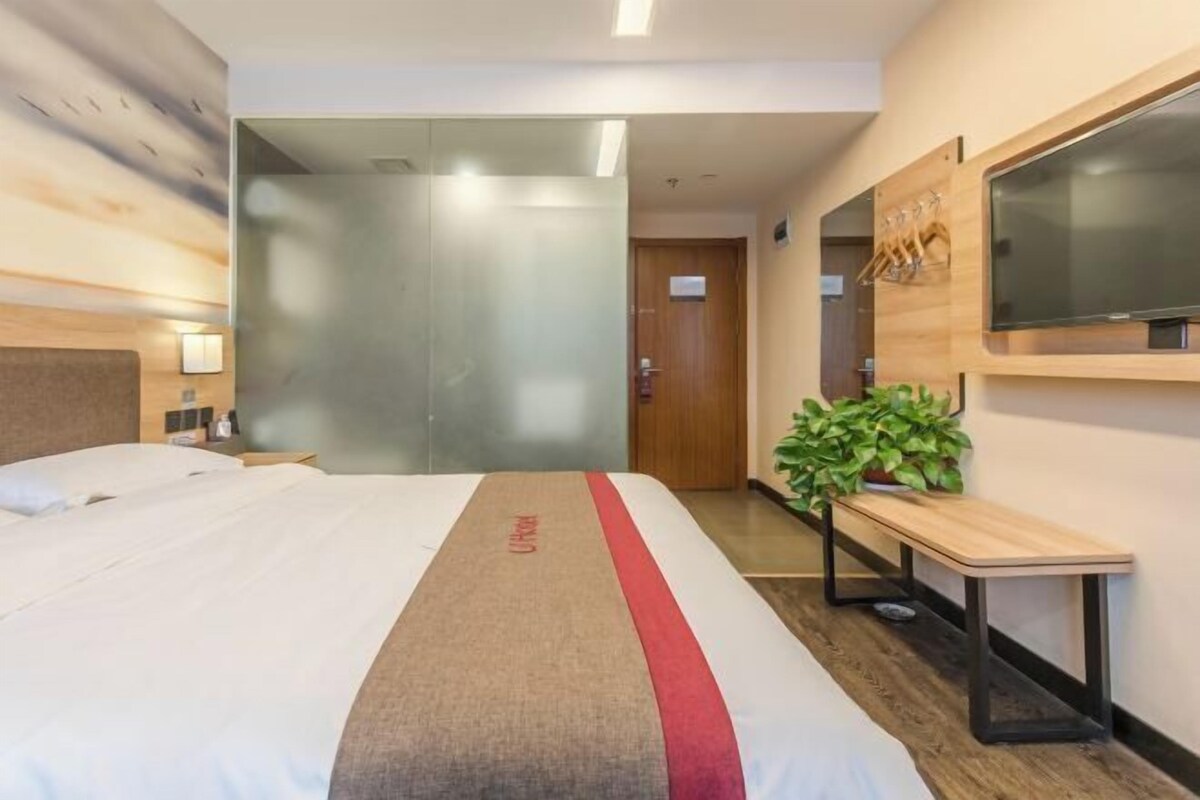 Quarto de hotel com uma cama, televisão presa à parede, um banco de madeira e um vaso com planta.