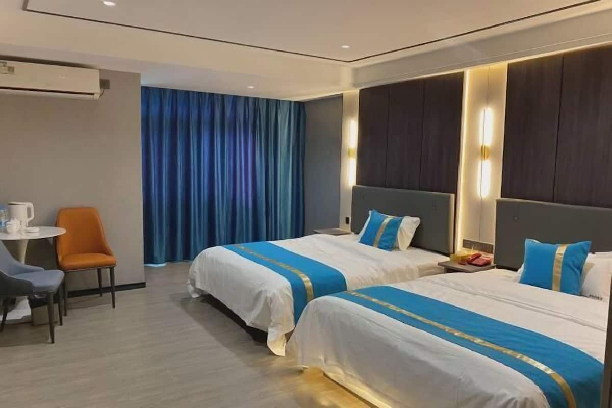 Quarto de hotel com duas camas, uma mesinha com cadeiras, uma máquina de café e uma janela grande com cortinas azuis.
