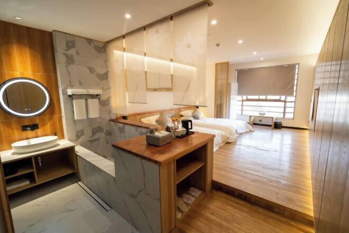 Quarto de hotel moderno com piso de madeira, espelho redondo, penteadeira de madeira, cama e uma janela grande.