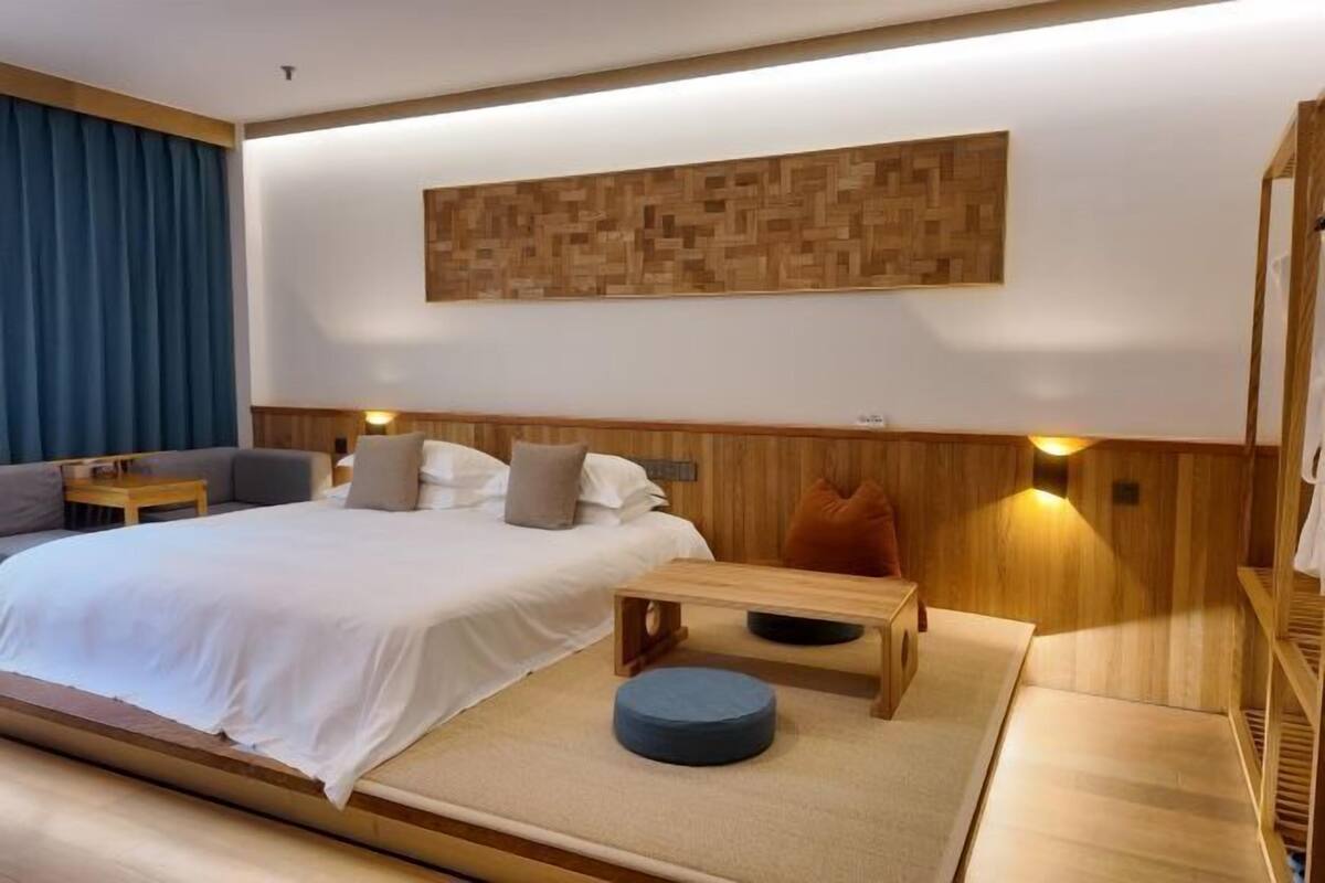 Quarto de hotel moderno com uma cama grande, cabeceira de madeira, uma obra de arte na parede e um pequeno espaço de estar com um banco e um pufe.