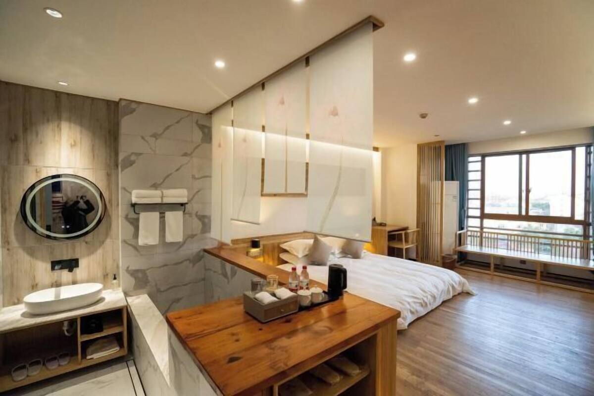 Quarto de hotel com toucador de madeira, espelho redondo, cama com roupa de cama branca, mesa de cabeceira com produtos de higiene e uma grande janela com vista.