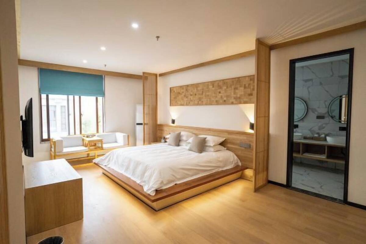 Quarto de hotel moderno com uma cama grande, uma escrivaninha, uma cadeira e banheiro com chuveiro de vidro.