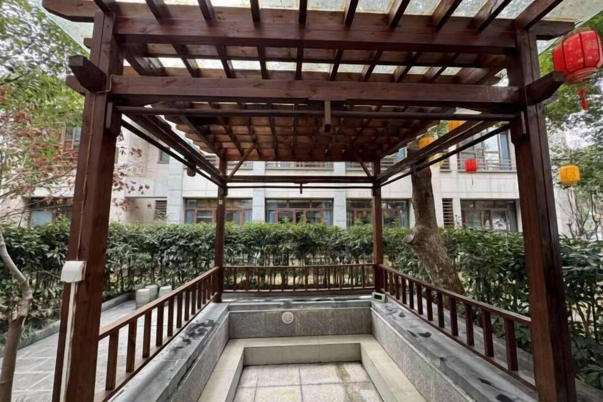 Uma estrutura de pérgola de madeira com um banco de pedra, rodeada por vegetação e edifícios ao fundo.