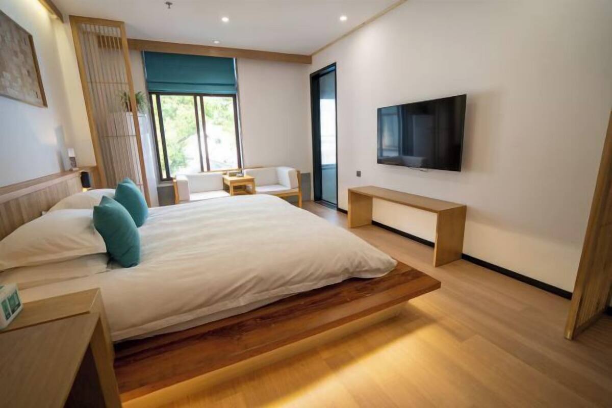 Quarto de hotel moderno com uma cama grande, televisão na parede, área de estar com um banco e vista para o verde lá fora.