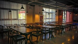 Uma ampla sala de jantar com várias mesas e cadeiras de madeira, janelas grandes com cortinas leves e luminárias pendentes.