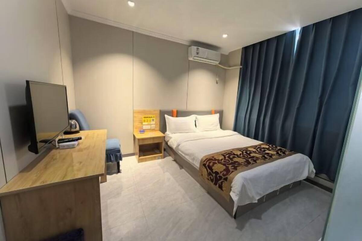 Quarto de hotel com cama, escrivaninha e cadeira, televisão, mesa de cabeceira com abajur, e janela com cortinas.