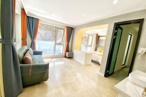Quarto de hotel moderno com sofá, uma pequena mesa de escritório, banheiro com pia e grandes janelas com cortinas.