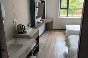 Quarto de hotel pequeno com uma cama, mesa de cabeceira com chaleira, televisão e janela com vista para o verde.