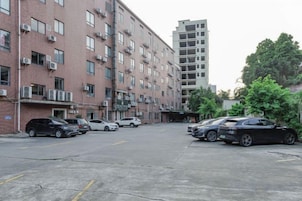 Um estacionamento com vários carros, um edifício de tijolos com várias andares e unidades de ar condicionado, e um prédio mais alto ao fundo.