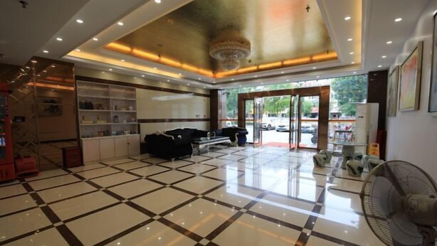 Um saguão de hotel com piso quadriculado, balcão de recepção, sofá preto e um ventilador.
