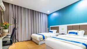 Quarto de hotel com duas camas, uma escrivaninha, uma cadeira, uma planta em vaso e uma cortina.