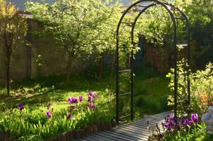 Um jardim com um caminho de madeira, um arco de metal e diversas plantas e flores.
