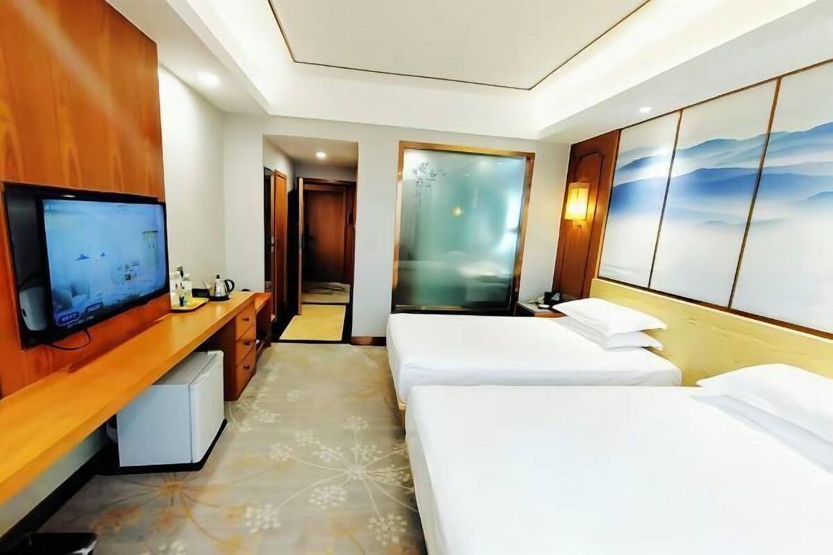 Quarto de hotel com duas camas, uma televisão sobre um console de madeira, uma pequena cozinha e uma janela grande com vista.