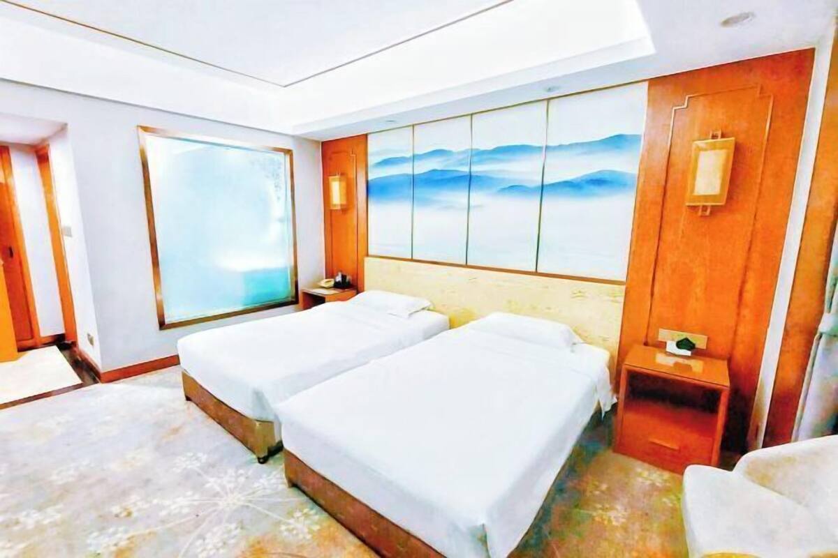 Quarto de hotel com duas camas, uma janela grande, um quadro na parede e uma mesa pequena com um abajur.